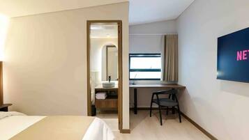 Quarto de hotel com cama, escrivaninha, cadeira, televisão e banheiro visível por uma porta aberta.