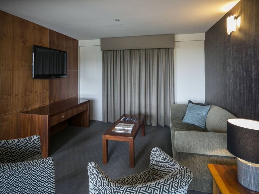 Quarto de hotel moderno com parede revestida em madeira, televisão de tela plana, sofá, poltrona estampada, mesa de centro e luminária.