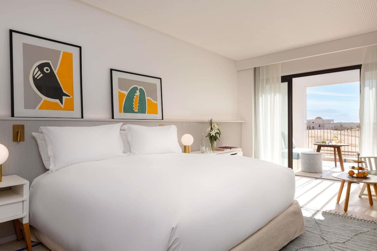 Quarto de hotel com uma cama grande, uma mesa de cabeceira com luminária, uma mesinha com um vaso e livros, e uma varanda com vista.