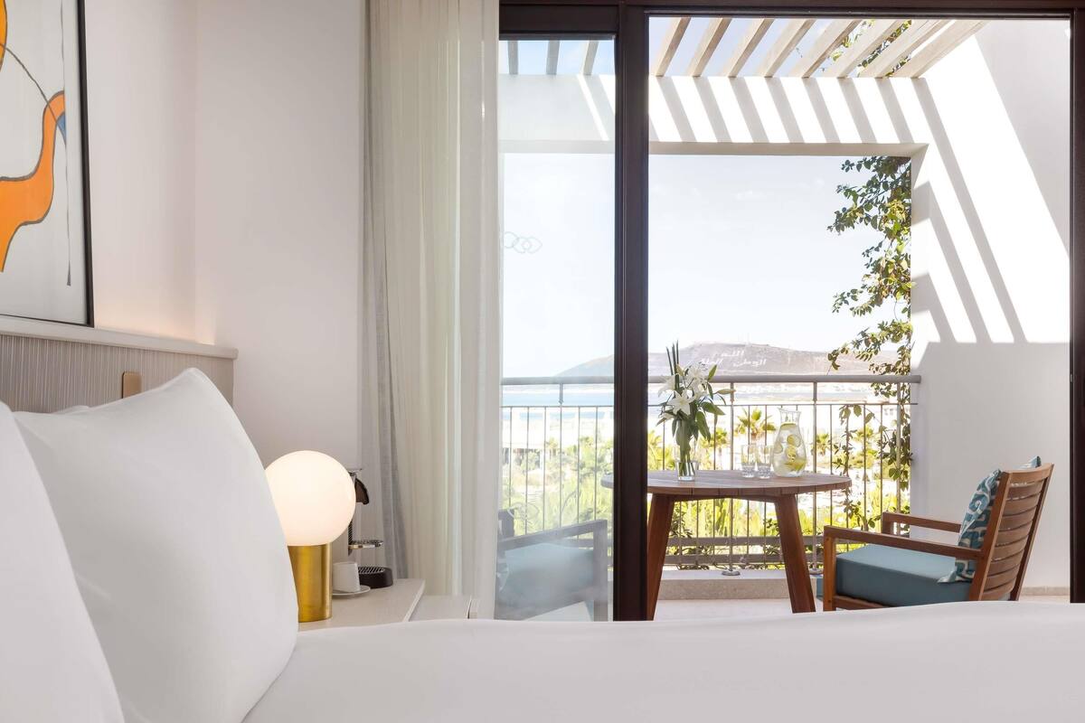 Quarto de hotel com cama, mesa de cabeceira com abajur, vista para a varanda com mesa e cadeiras, e um quadro na parede.
