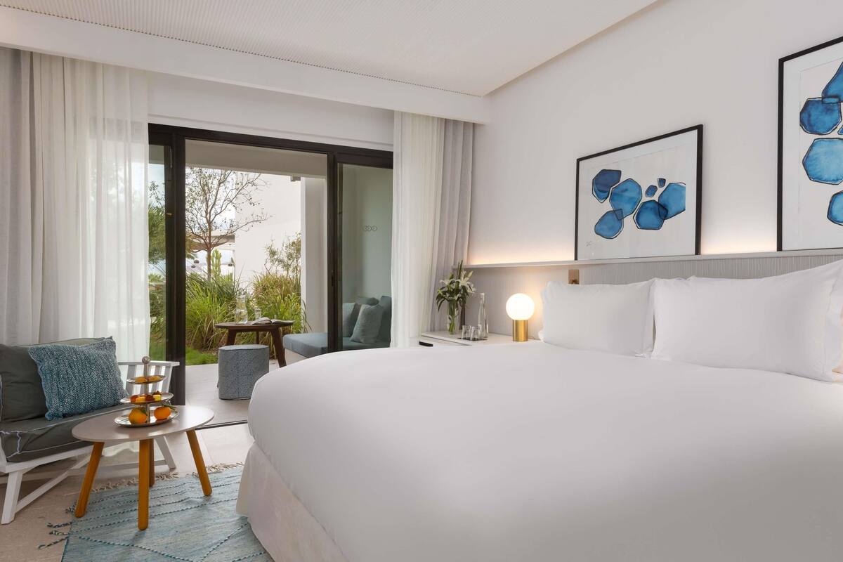 Quarto de hotel com uma cama grande, uma mesinha com abajur, uma cadeira e vista para o exterior.