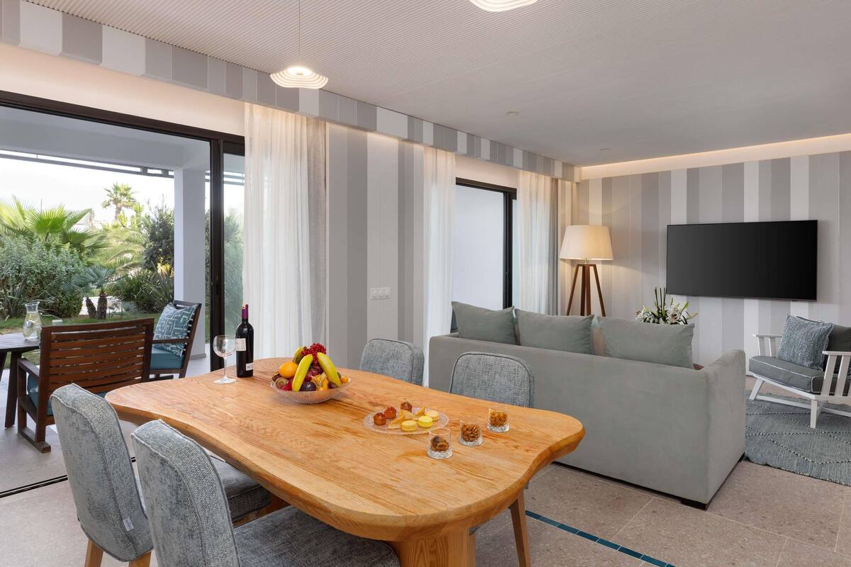 Uma sala de estar moderna com uma mesa de jantar de madeira, cadeiras estofadas cinzas, um sofá cinza e uma TV de tela plana fixada na parede.