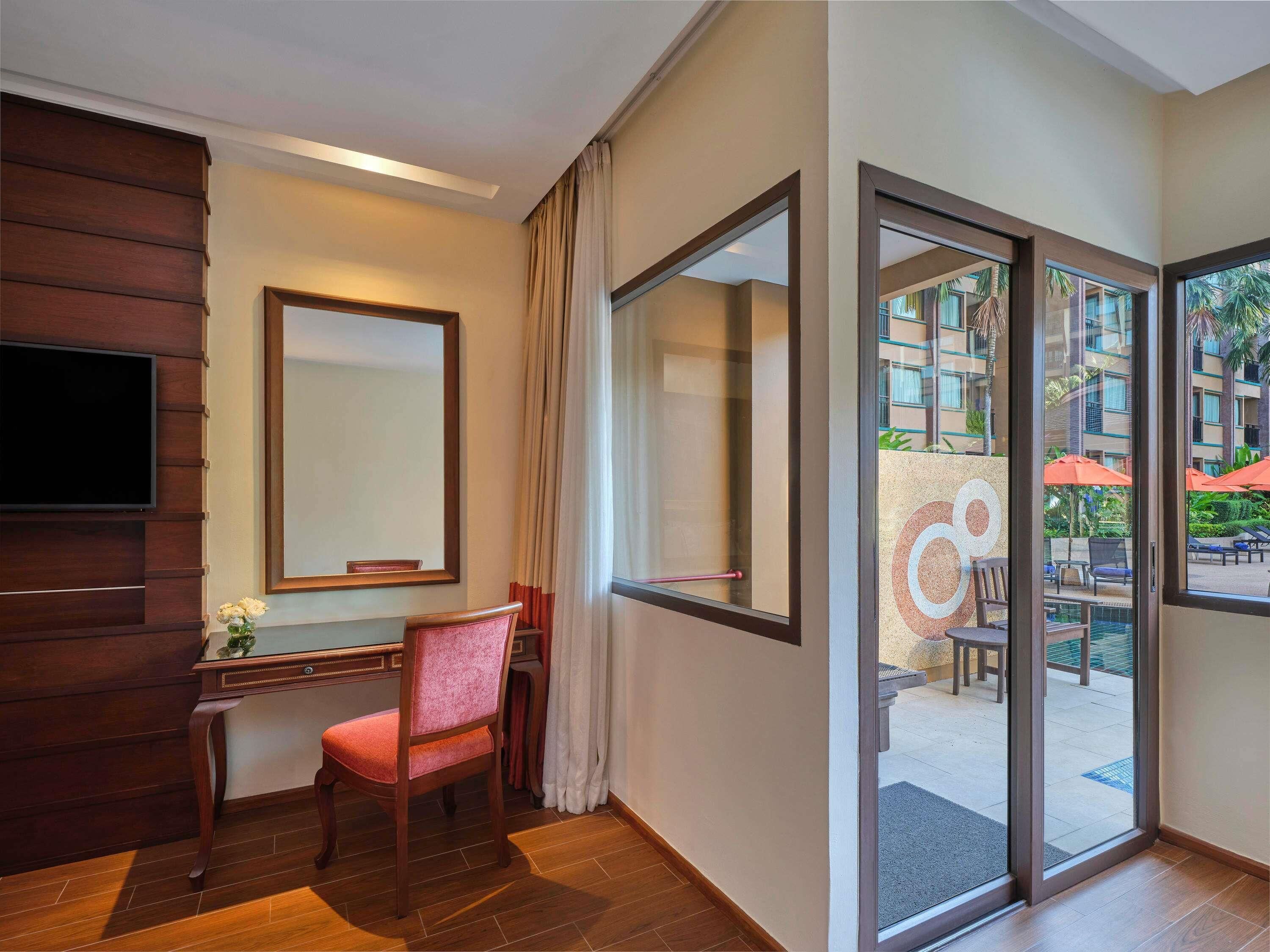 Quarto de hotel com escrivaninha de madeira, uma cadeira, um espelho grande e uma porta de vidro deslizante para área externa com guarda-sóis e assentos.