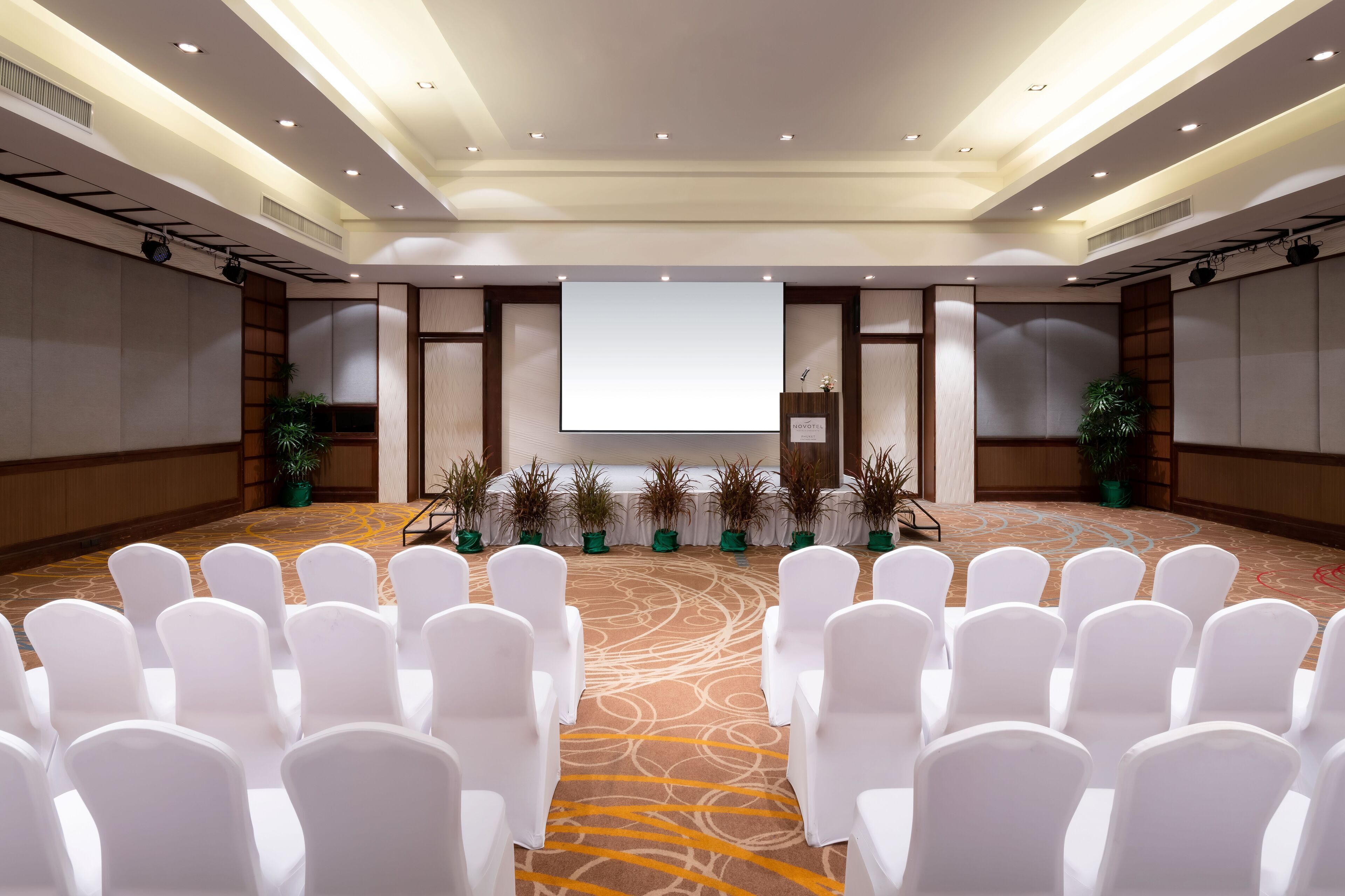 Sala de conferência com fileiras de cadeiras brancas, uma tela grande e plantas em vasos.