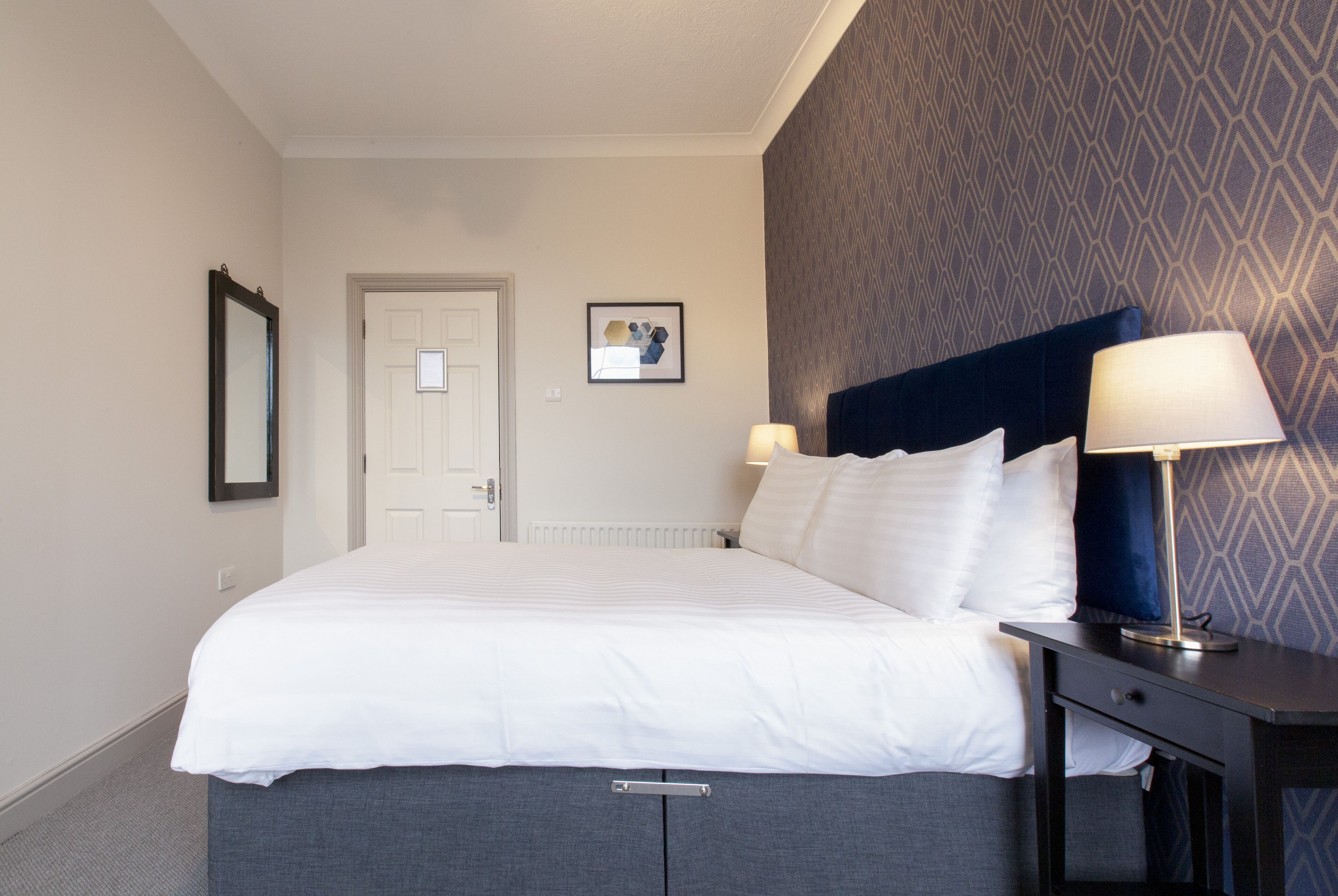 Quarto de hotel com cama, mesas de cabeceira, painel de cabeceira, porta e parede estampada.