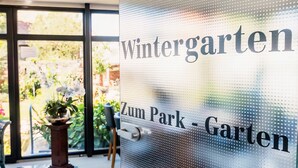 Placa com a inscrição 'Wintergarten Zum Park - Garten', com vista para plantas em vasos e uma porta.