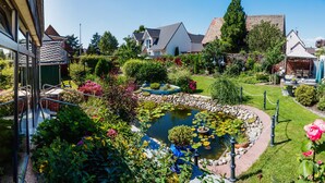 Um jardim com lago, diversas plantas e um edifício com varanda.