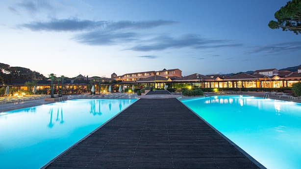 Piscina com um deck de madeira que a conduz, rodeada por edifícios com iluminação quente.
