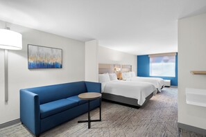 Quarto de hotel com sofá azul, mesa redonda, cama com roupa de cama branca e um quadro na parede.
