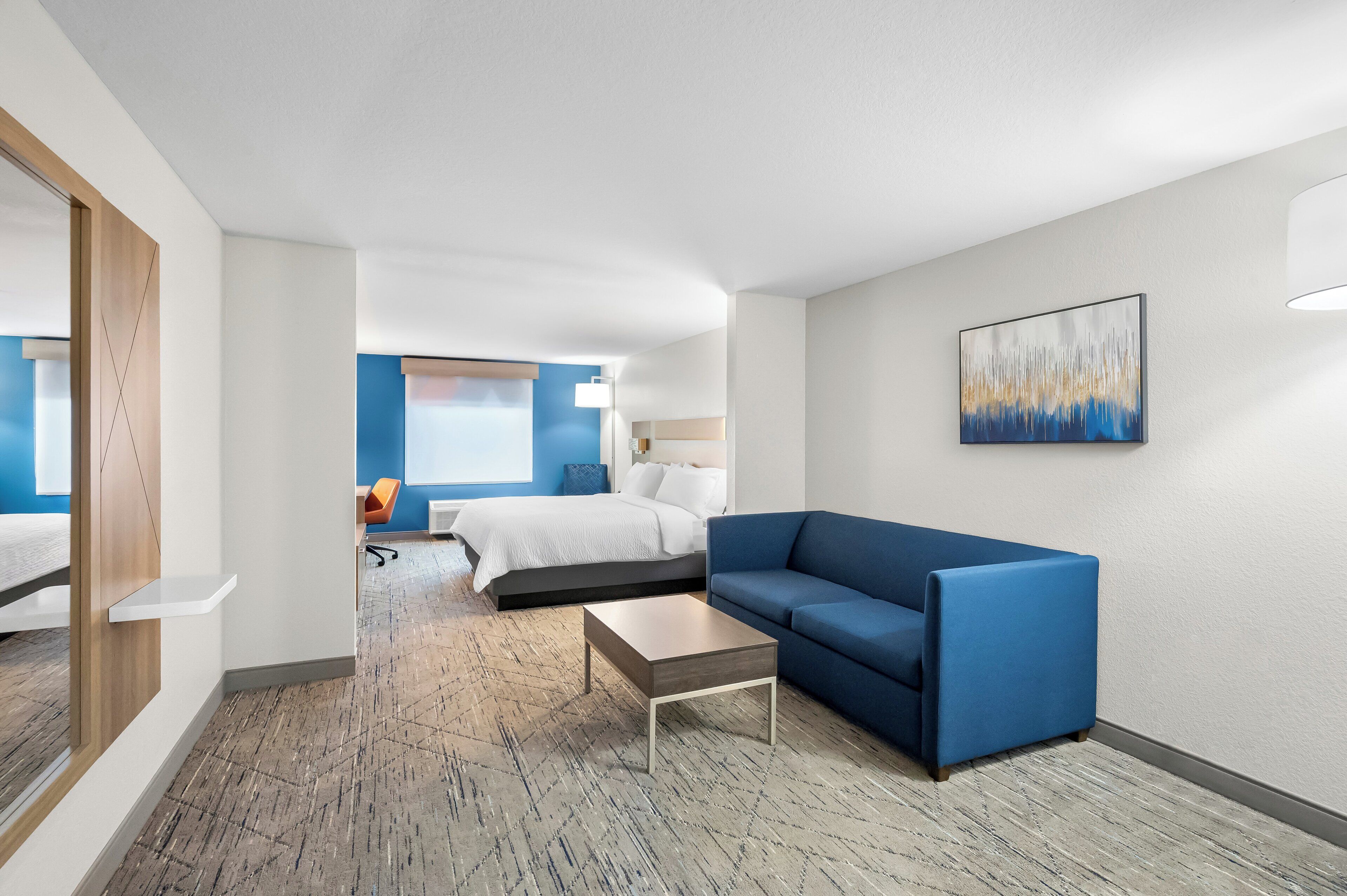 Quarto de hotel moderno com uma cama, um sofá azul, uma mesinha e um quadro na parede.