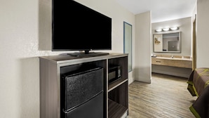 Quarto de hotel com TV de tela plana em suporte de madeira, micro-ondas, mini-geladeira, banheiro com pia e espelho, e cama com manta marrom.