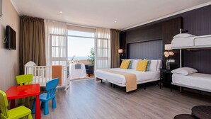 Quarto de hotel com uma cama grande, uma cama menor, uma escrivaninha com cadeira, uma mesa de jantar com cadeiras e vista para o exterior.