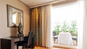 Quarto de hotel com escrivaninha, cadeira e uma janela grande com cortinas.