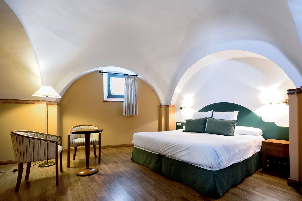 Quarto de hotel com teto arredondado, cama com roupa de cama verde, uma mesinha e uma cadeira, e janela com cortinas.