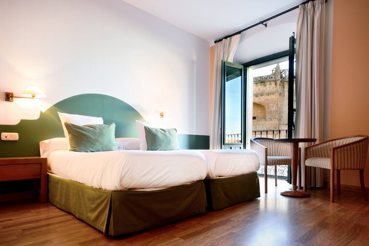 Quarto de hotel com uma cama grande, uma mesinha e vista para um edifício histórico pela janela.