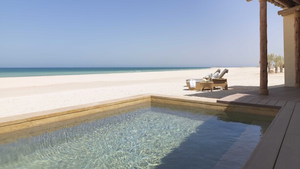 Uma piscina com água cristalina, uma praia e uma área de descanso com cadeiras e uma mesa.