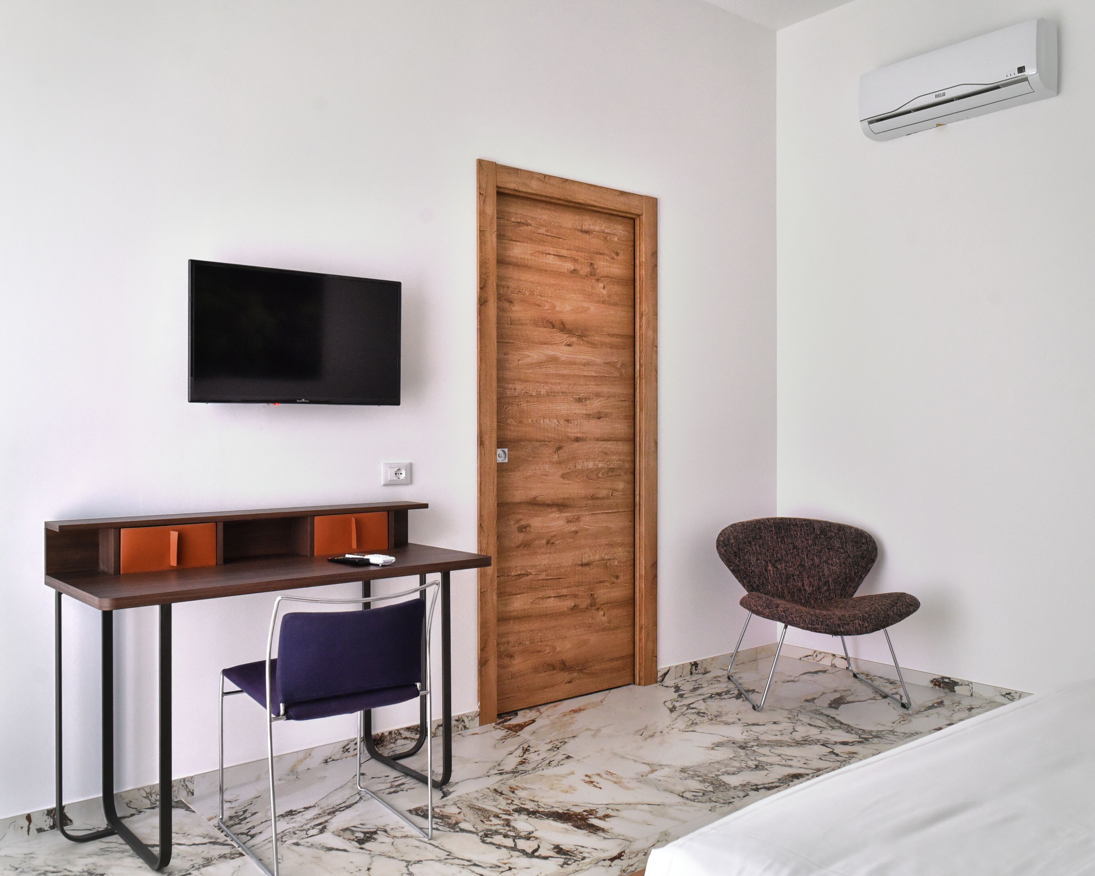 Quarto de hotel com porta de madeira, televisão na parede, secretária com cadeira e unidade de ar condicionado.