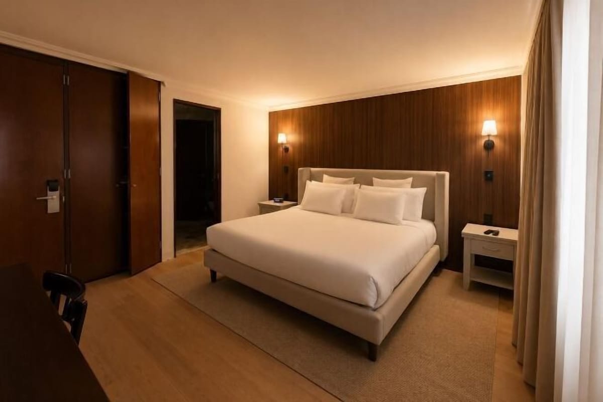Quarto de hotel com uma cama grande, mesas de cabeceira, um guarda-roupa de madeira e chão coberto de carpete.