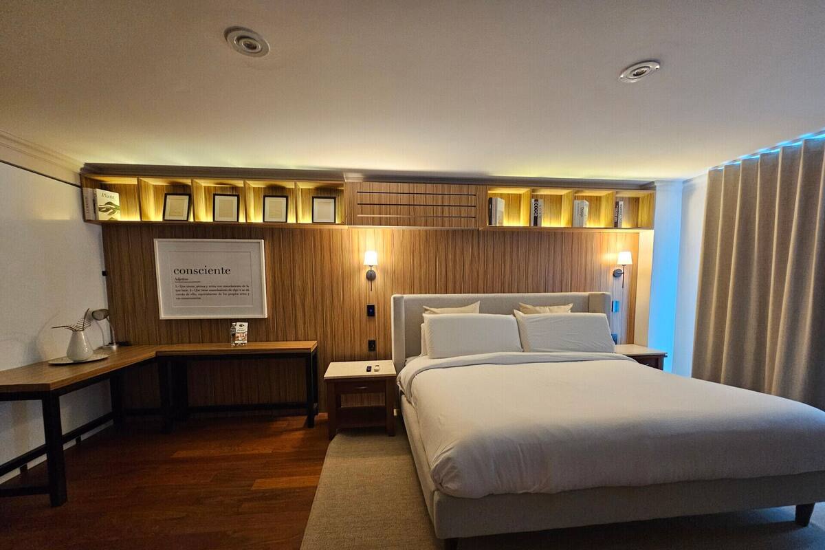 Quarto de hotel com uma cama grande, painéis de madeira, uma escrivaninha com abajur e um quadro na parede.
