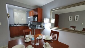 Uma sala de jantar com uma mesa de madeira posta para quatro pessoas, uma cozinha com micro-ondas e armários, e um espelho grande na parede.