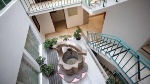 Um edifício com vários níveis e um pátio central com uma fonte de pedra, plantas em vasos e uma escadaria com grades azuis.