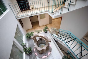Um edifício com vários níveis e um pátio central com uma fonte de pedra, plantas em vasos e uma escadaria com grades azuis.