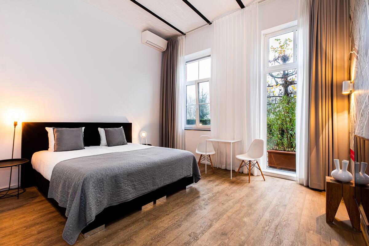 Quarto de hotel moderno com uma cama grande, mesas de cabeceira, uma pequena escrivaninha e uma janela com cortinas.