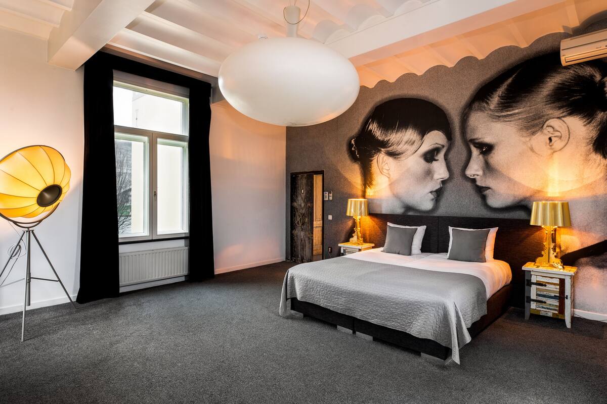 Quarto moderno com uma cama grande, duas mesas de cabeceira com abajures, uma janela ampla e um mural exclusivo na parede.