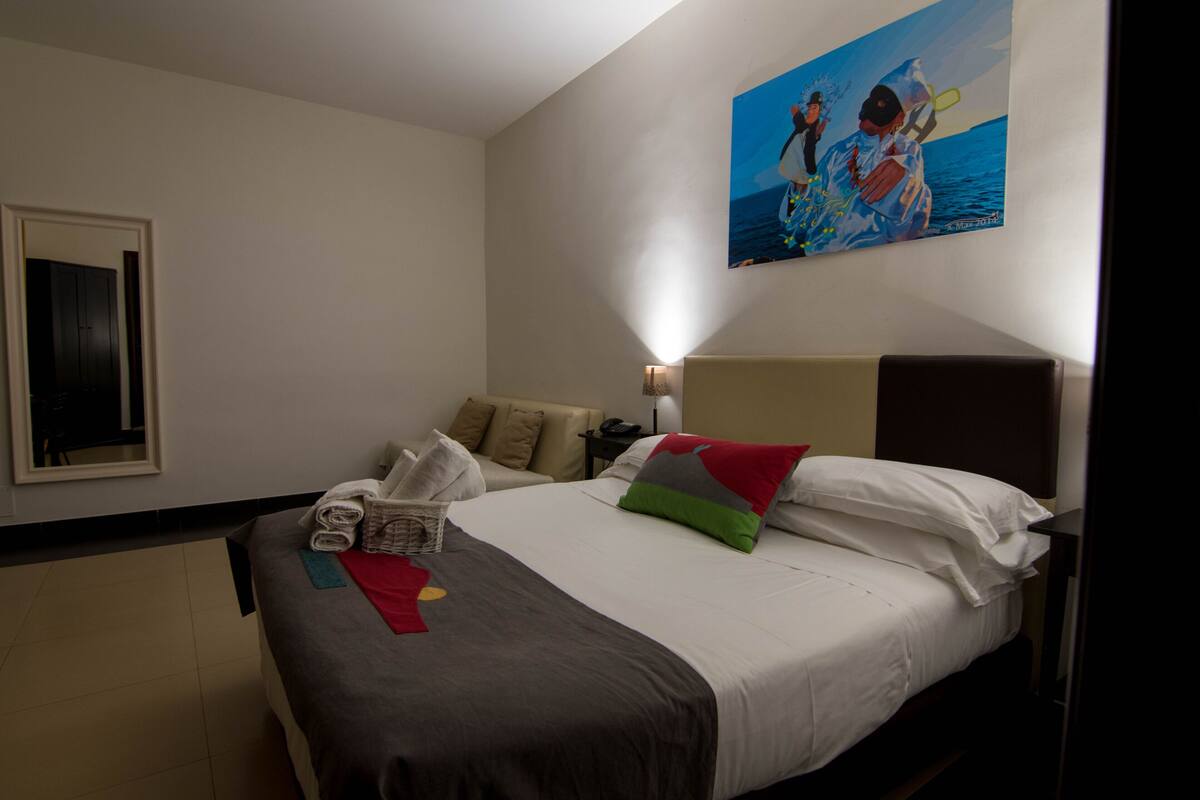 Quarto de hotel com cama, almofadas, mesa de cabeceira, abajur e um quadro na parede.