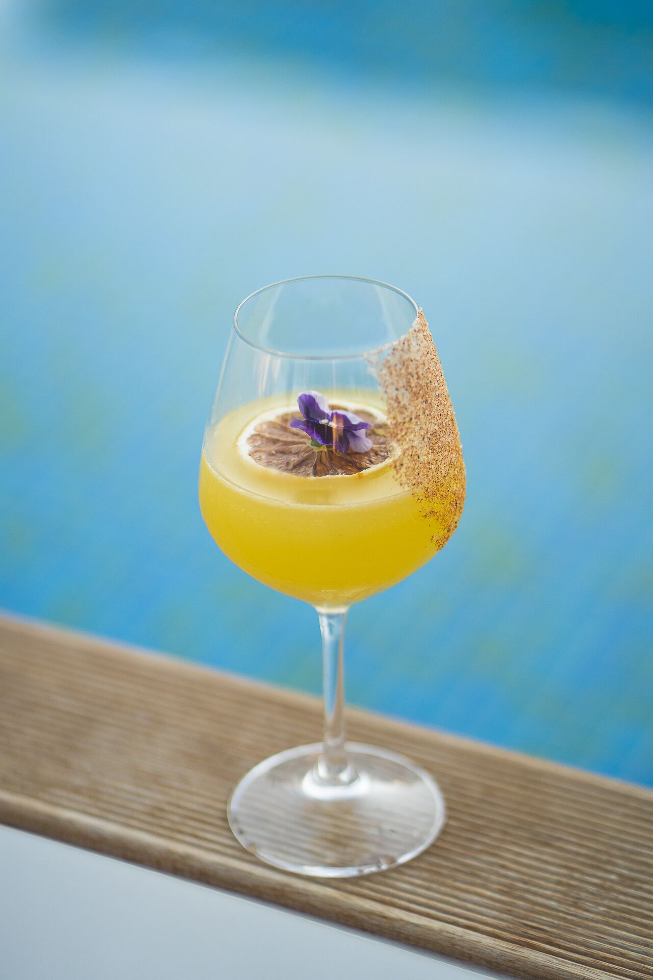 Um cocktail num copo de haste, com uma fatia de limão e uma flor roxa como enfeite, apoiado em uma superfície de madeira com um fundo azul desfocado.