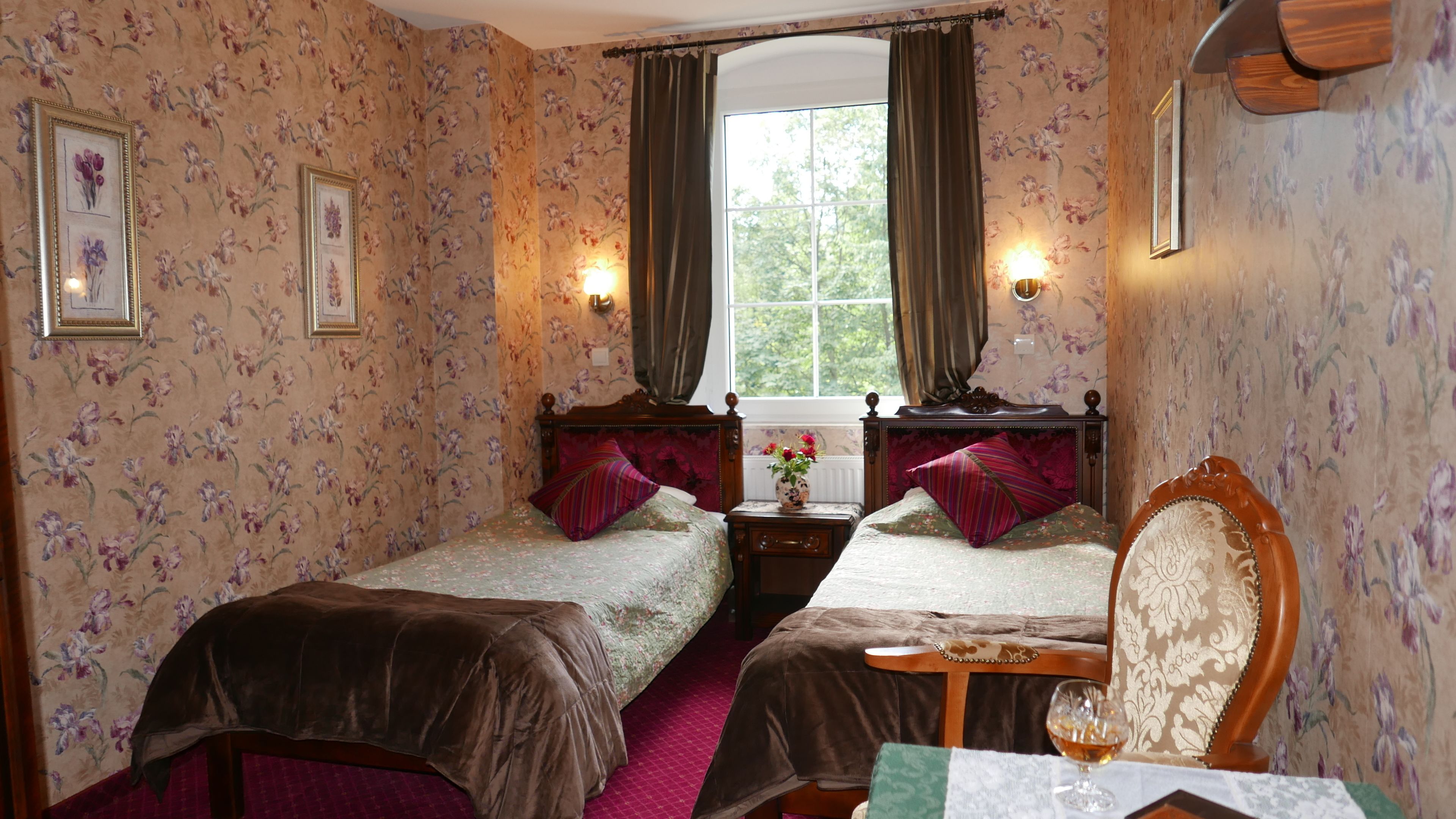 Quarto com duas camas, papel de parede floral, janela com cortinas e mesa de cabeceira com um vaso de flores.