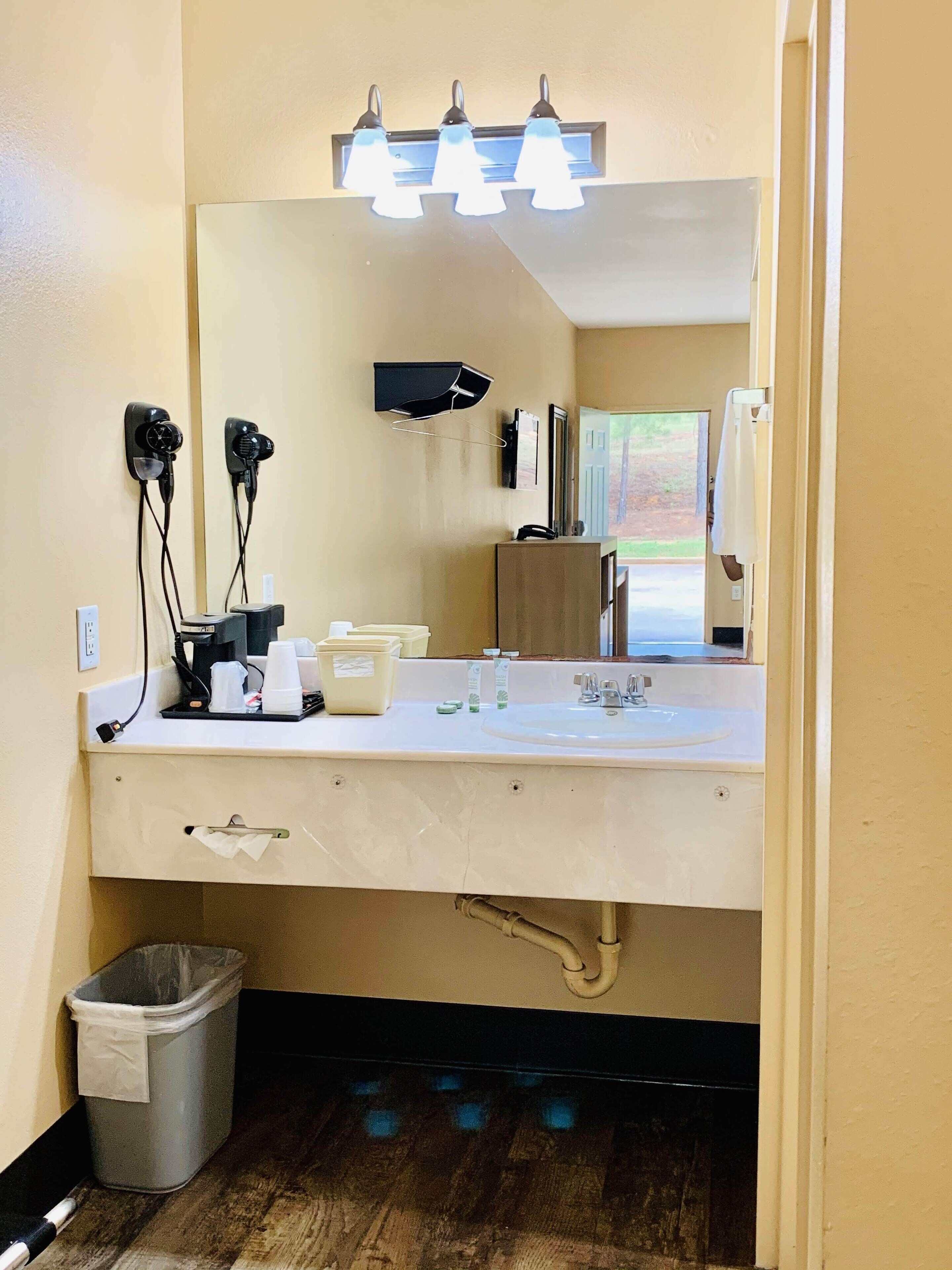 Casa de banho com lavatório, espelho e três luzes. Inclui secadores de cabelo e um contentor de lixo.