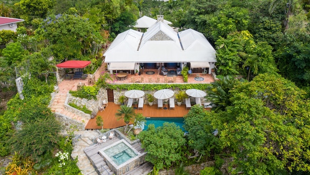 Uma grande villa com telhado branco e piscina, rodeada por vegetação exuberante e vista para o oceano.