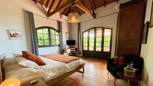 Quarto com cama, escrivaninha, cadeira e uma janela ampla com vista para área verde.