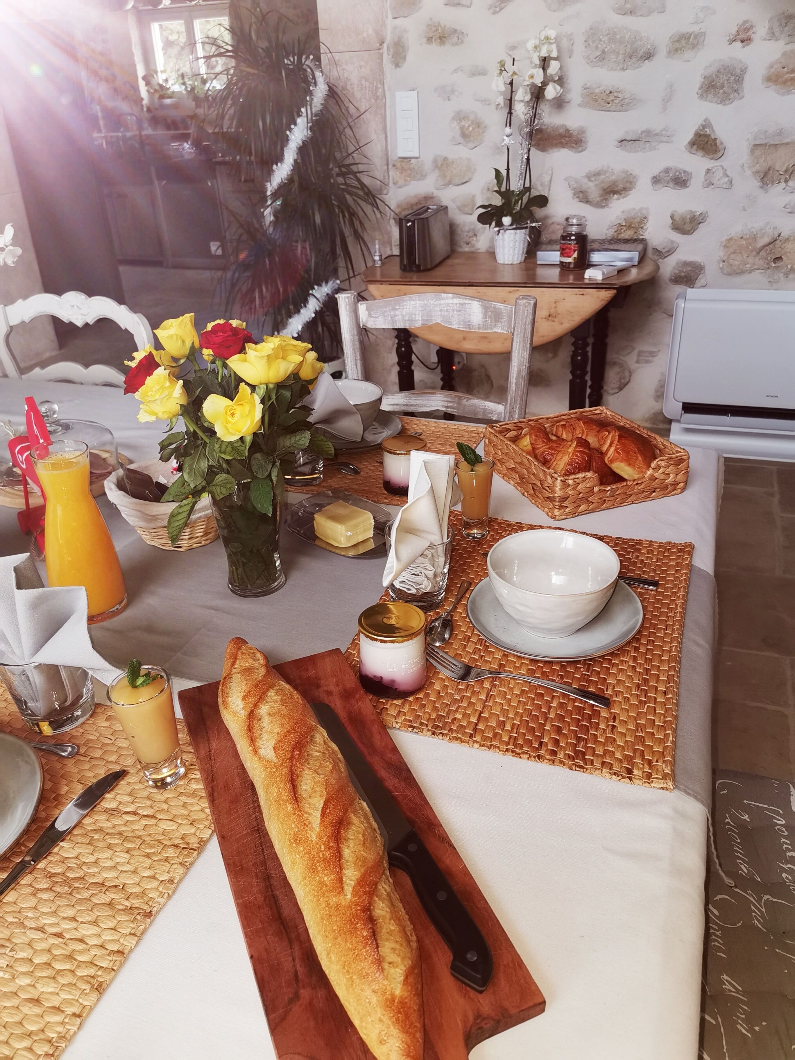 Mesa posta para dois, com pão, cesta de frutas, um vaso de flores e jarra de suco.