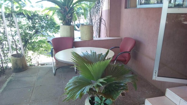 Uma área de pátio com uma mesa redonda e duas cadeiras vermelhas, rodeada por plantas em vasos.