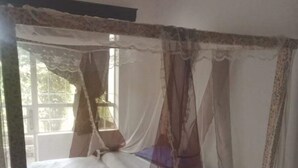 Quarto com cama coberta por mosquiteiro, janela com cortinas e chão de madeira.