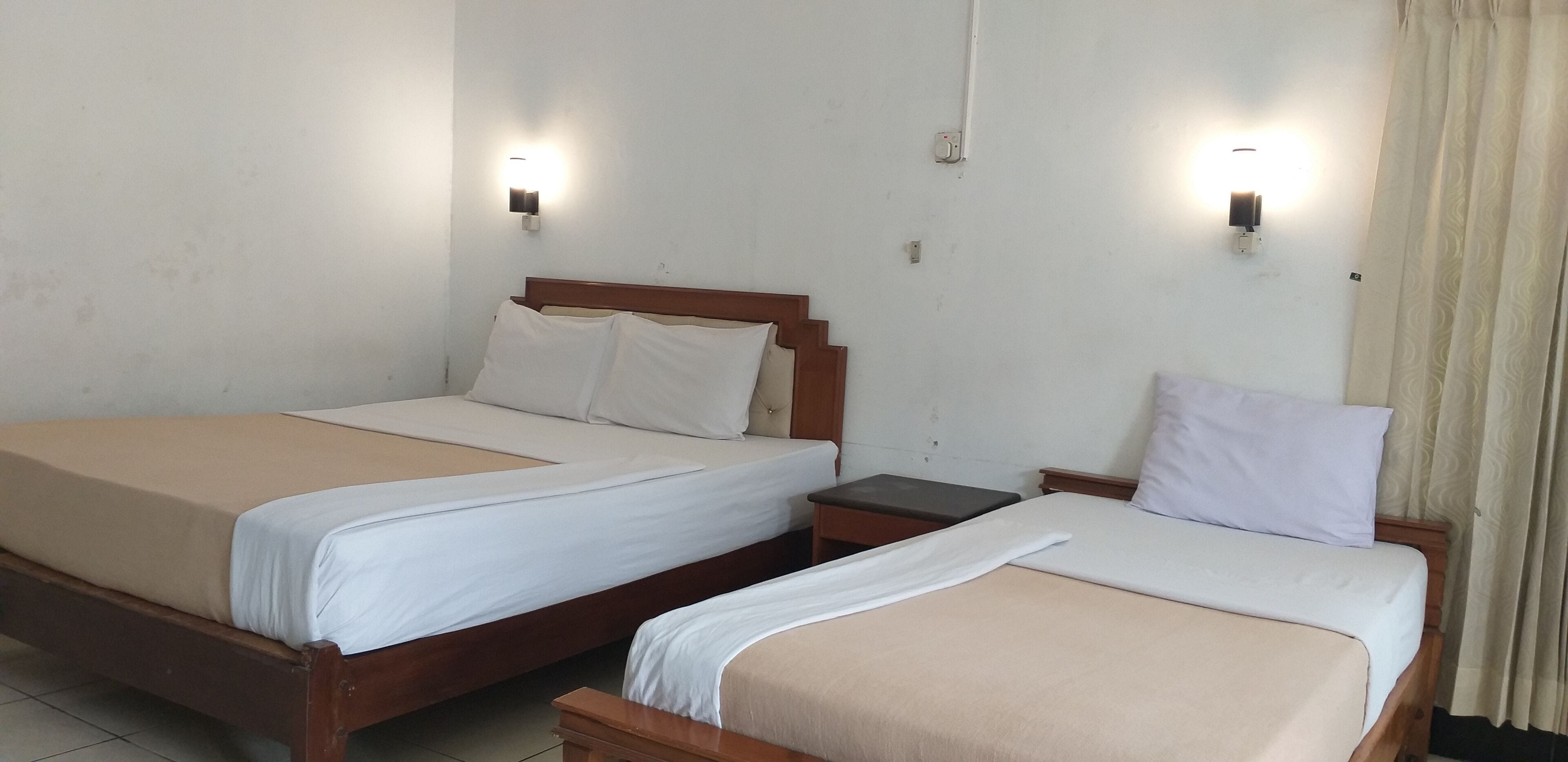 Duas camas de solteiro com estrutura de madeira e roupa de cama branca, uma pequena mesa de cabeceira e luminárias fixadas na parede.