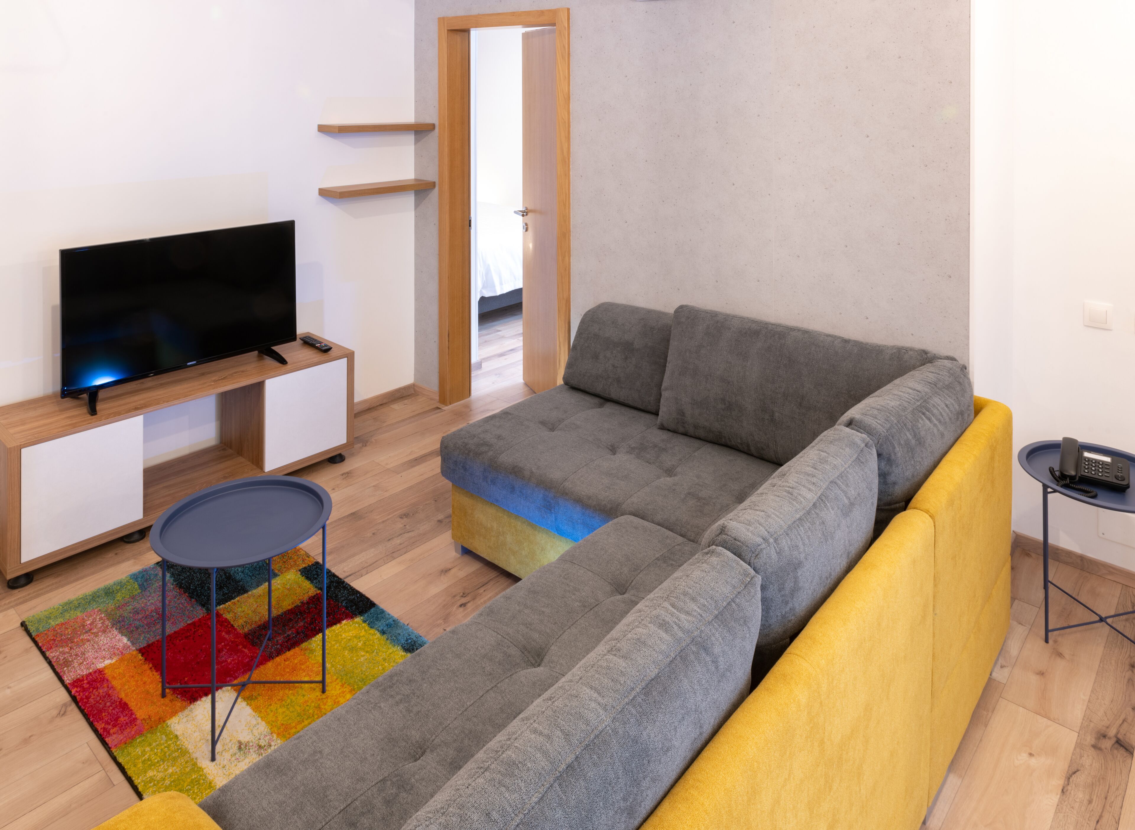 Uma sala de estar moderna com um sofá seccional cinza, uma mesa de centro redonda pequena, uma TV de tela plana em um suporte de madeira e um tapete colorido.