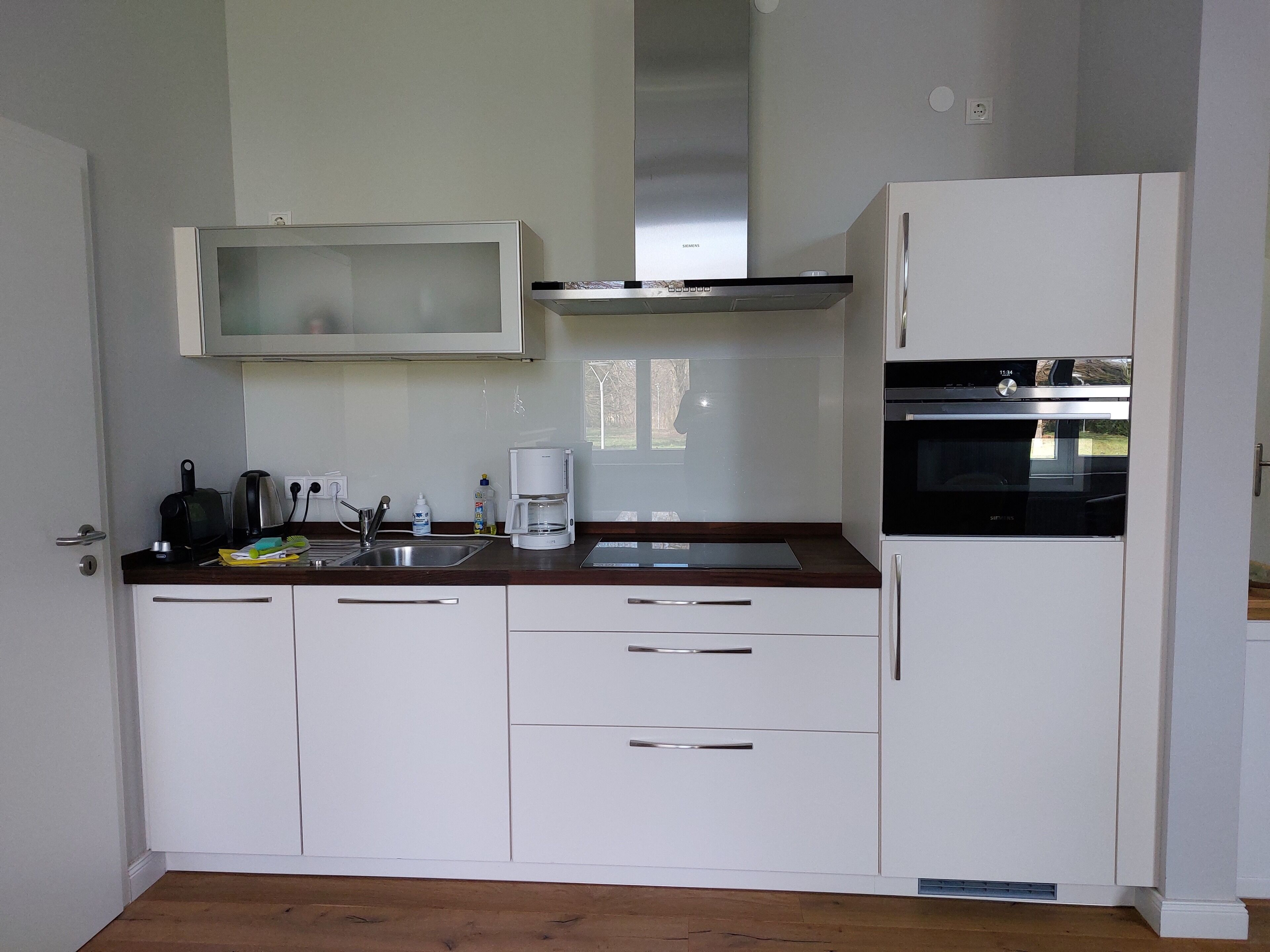Cozinha compacta com armários brancos, coifa de aço inoxidável, micro-ondas embutido, cafeteira e forno preto.