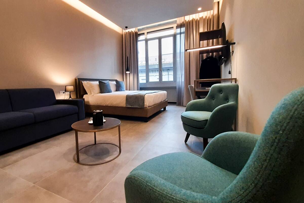 Quarto de hotel moderno com uma cama, um sofá, uma poltrona verde e uma pequena mesa de centro.