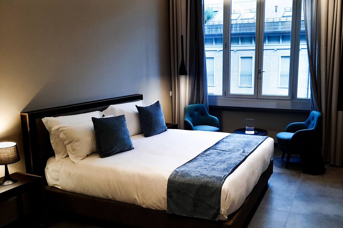 Quarto de hotel com cama, duas cadeiras, uma mesa pequena e uma janela com vista para edifícios.