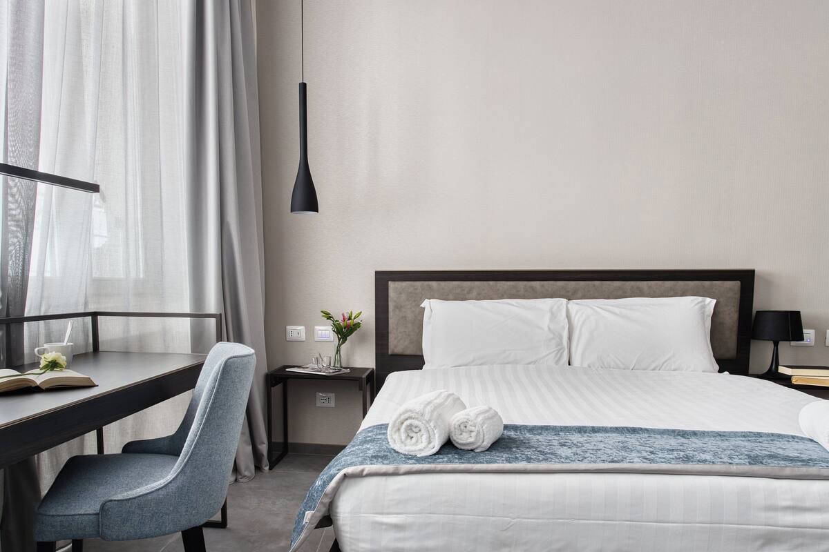 Quarto de hotel com cama, escrivaninha, cadeira, mesa de cabeceira e luminária.
