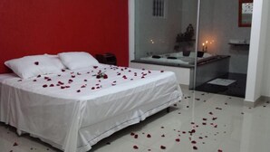 Quarto com cama coberta de pétalas de rosa, divisória de vidro, pequena mesa com velas e parede vermelha.