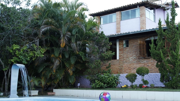 Uma piscina com cachoeira, rodeada por vegetação exuberante e uma casa moderna com fachada de tijolos.