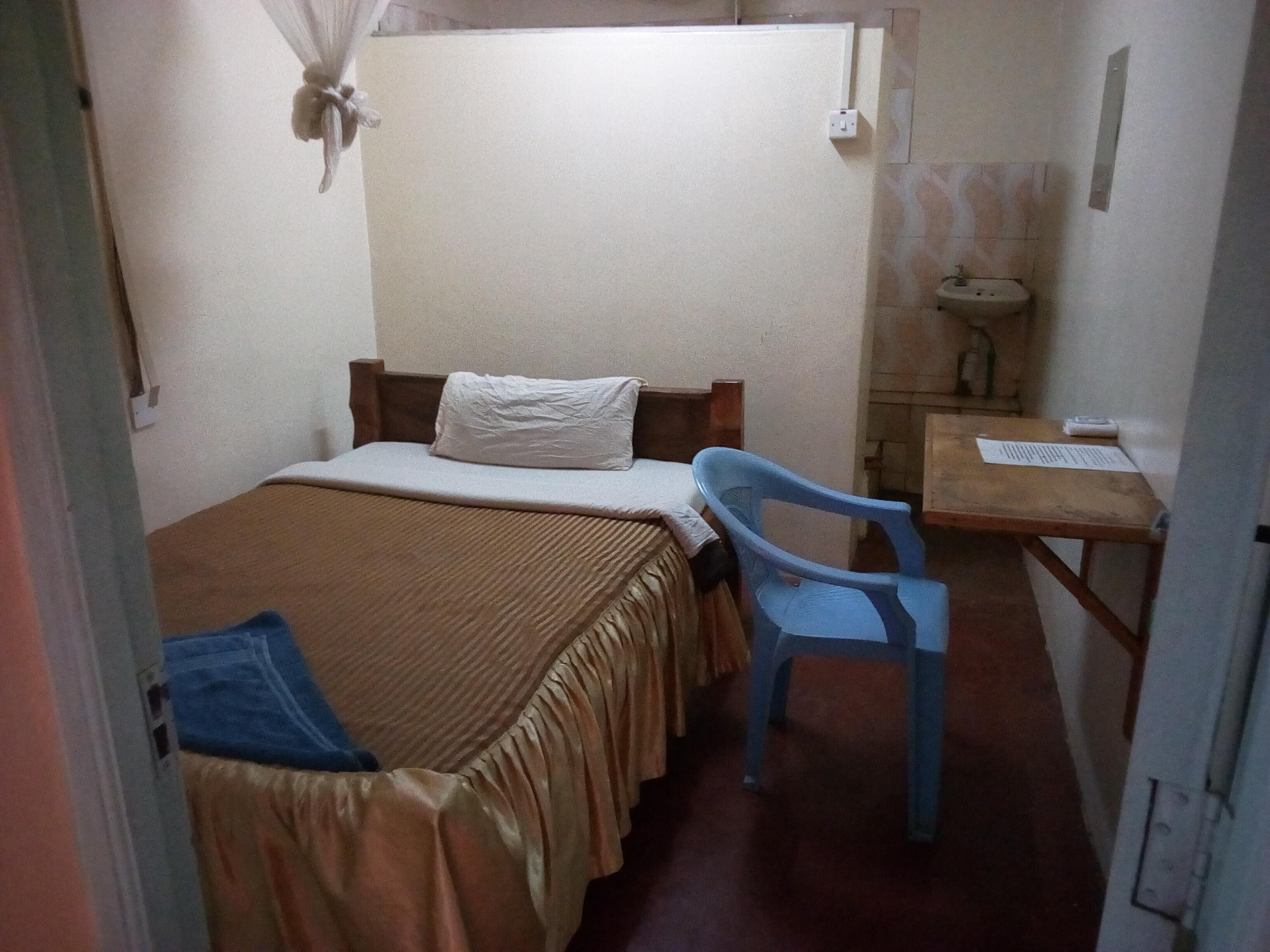 Quarto pequeno com uma cama de solteiro, cabeceira de madeira, mesa de cabeceira, uma cadeira e escrivaninha com pia.