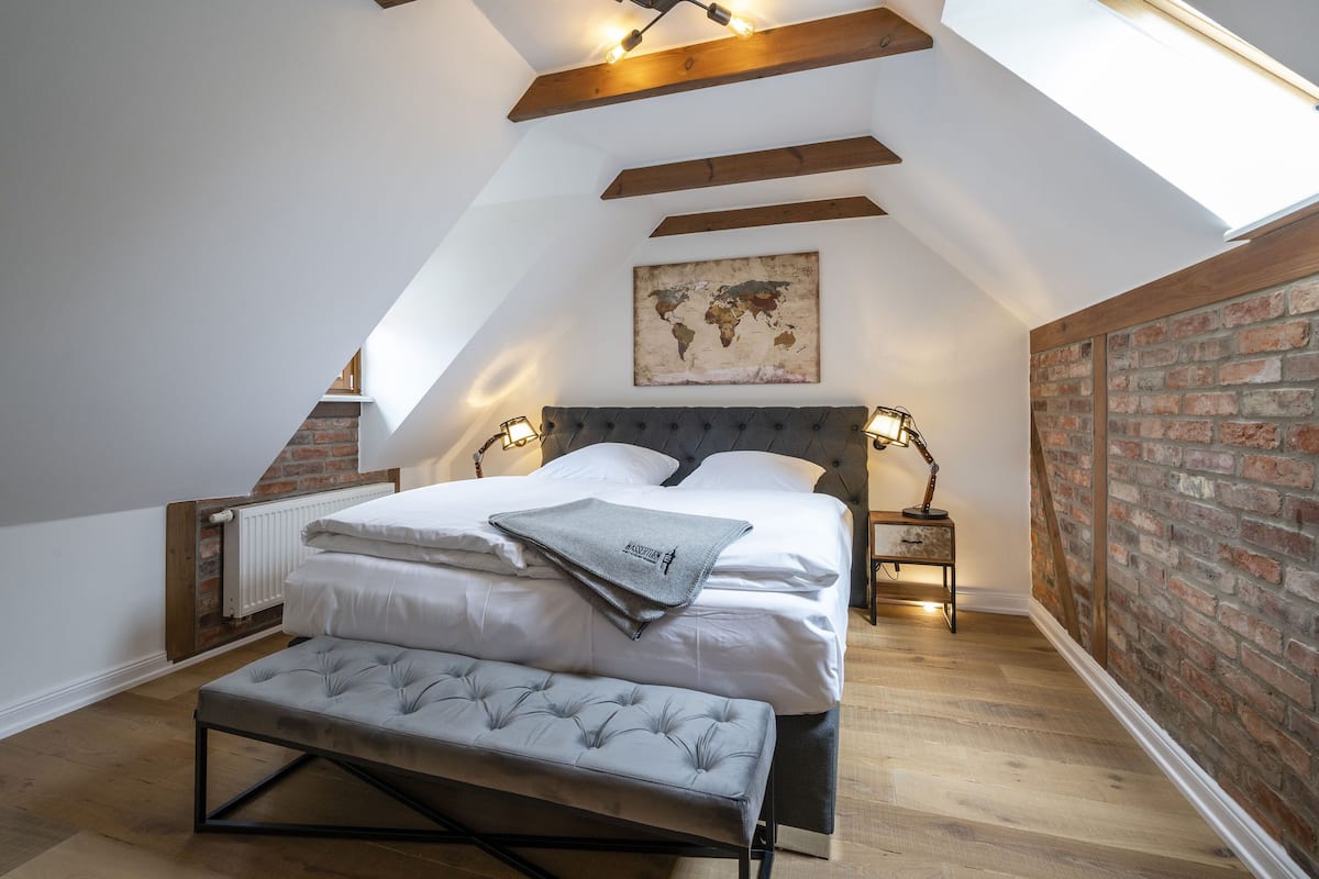 Um quarto em estilo loft com teto inclinado, vigas de madeira aparentes, um mapa-múndi na parede, uma cama com roupa de cama branca, duas mesas de cabeceira com abajures e um banco.