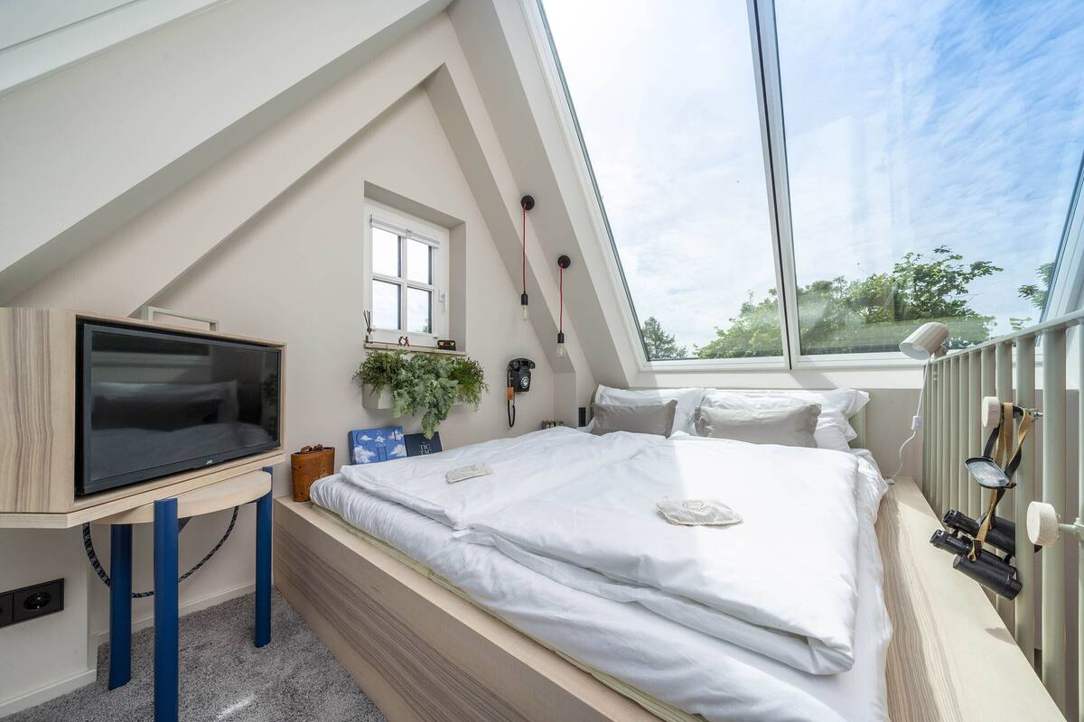 Quarto com televisão sobre suporte de madeira, cama com roupa de cama branca, janela com vista para árvores e uma pequena escrivaninha com uma planta.