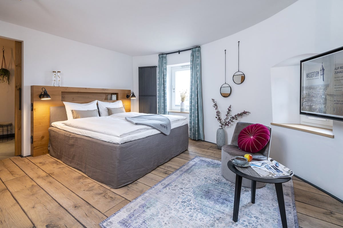 Quarto moderno com cama grande, mesa de cabeceira, uma pequena mesa com jornal e janela com cortinas.