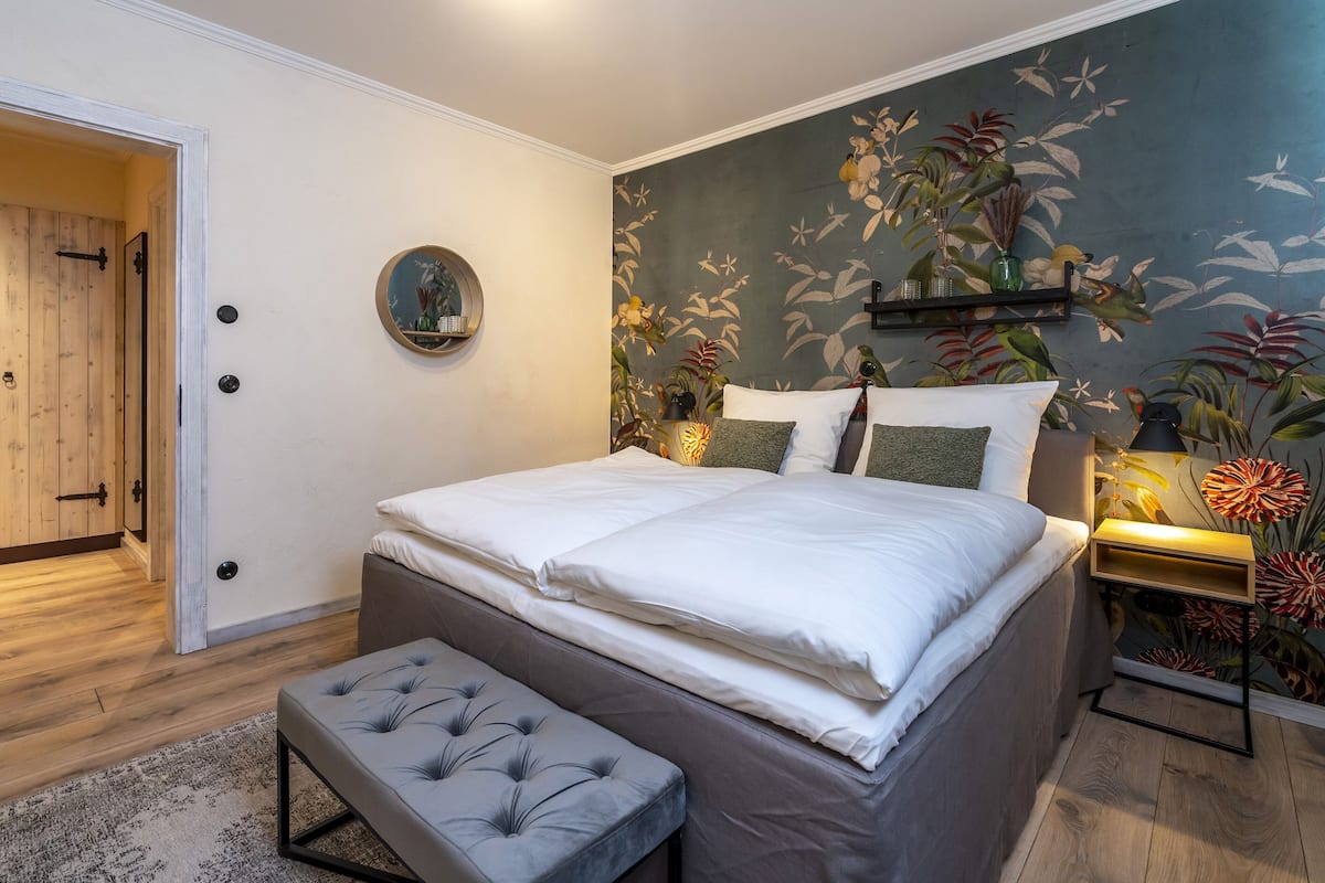 Um quarto com cama, um banco, mesa de cabeceira, prateleira na parede e uma porta.